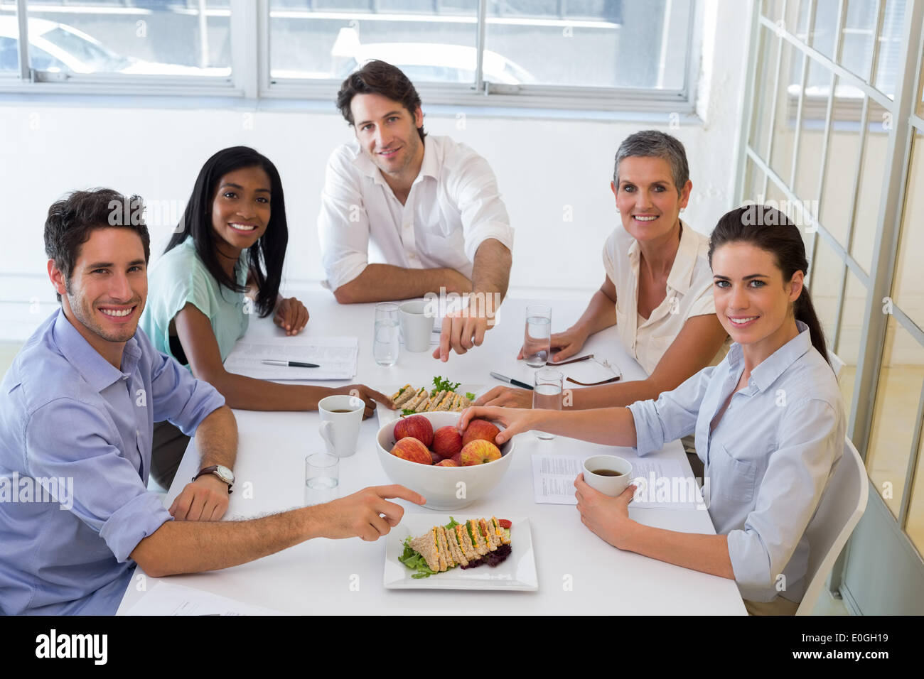 Business people smiling at camera eating sandwiches and fruit for lunch ...