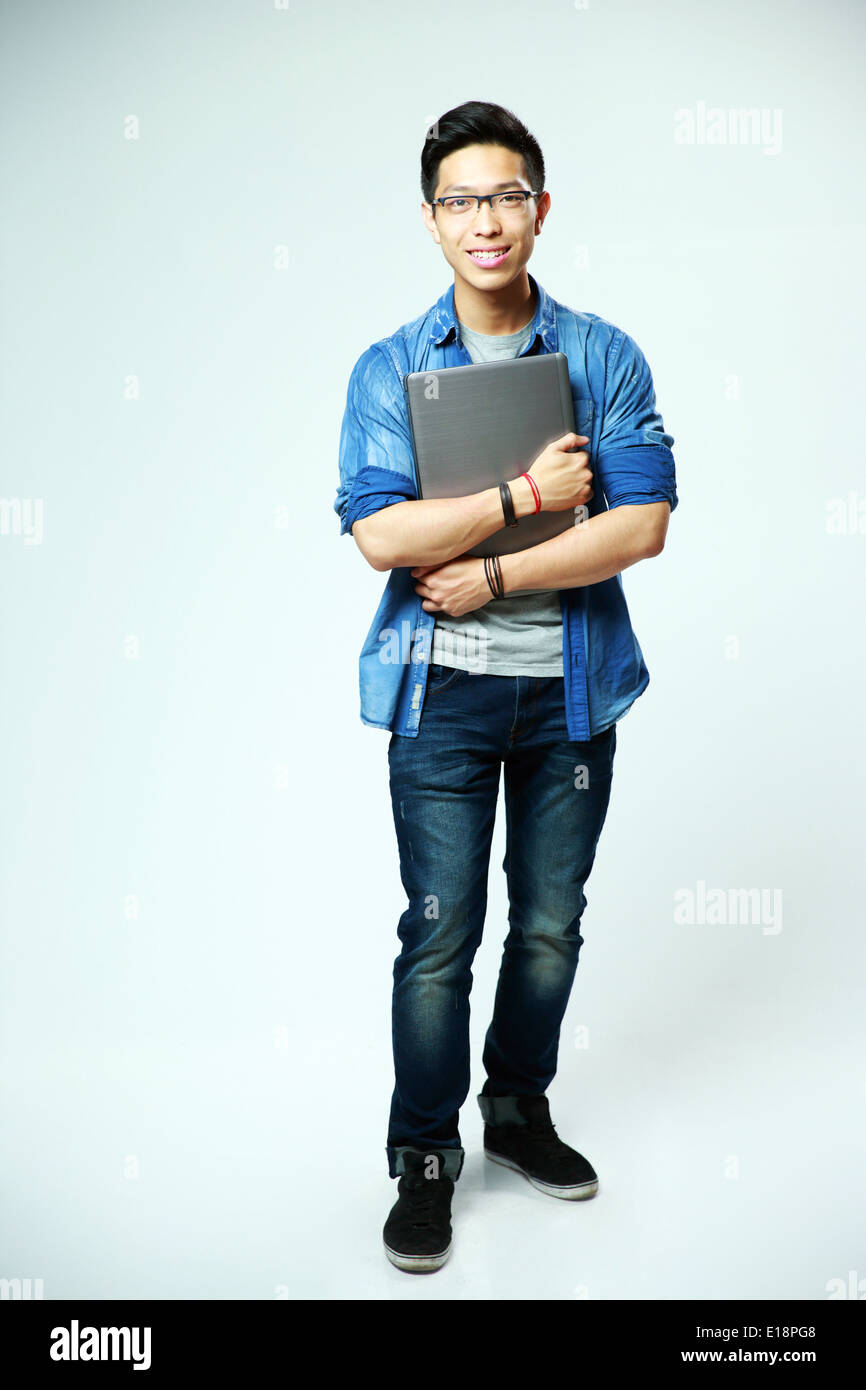 Full length portrait of a happy asian man standing with laptop on gray ...