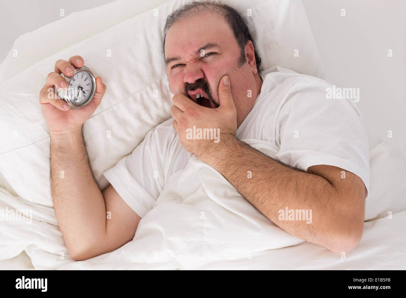 Man suffering from insomnia lying in bed clutching his alarm clock as ...