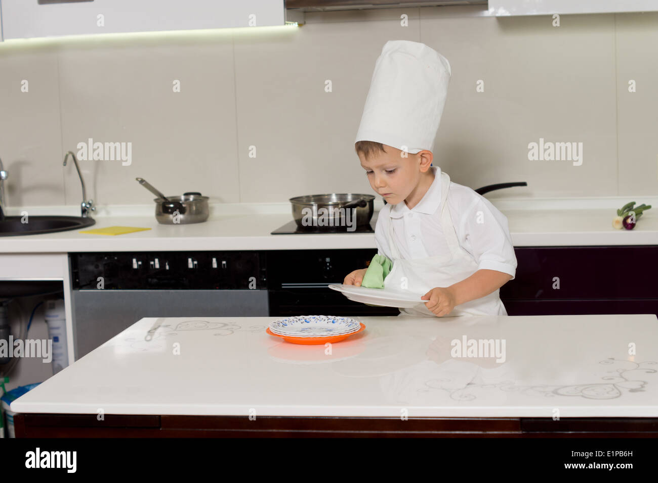 Small little boy in a chefs uniform wiping dishes and carefully placing ...