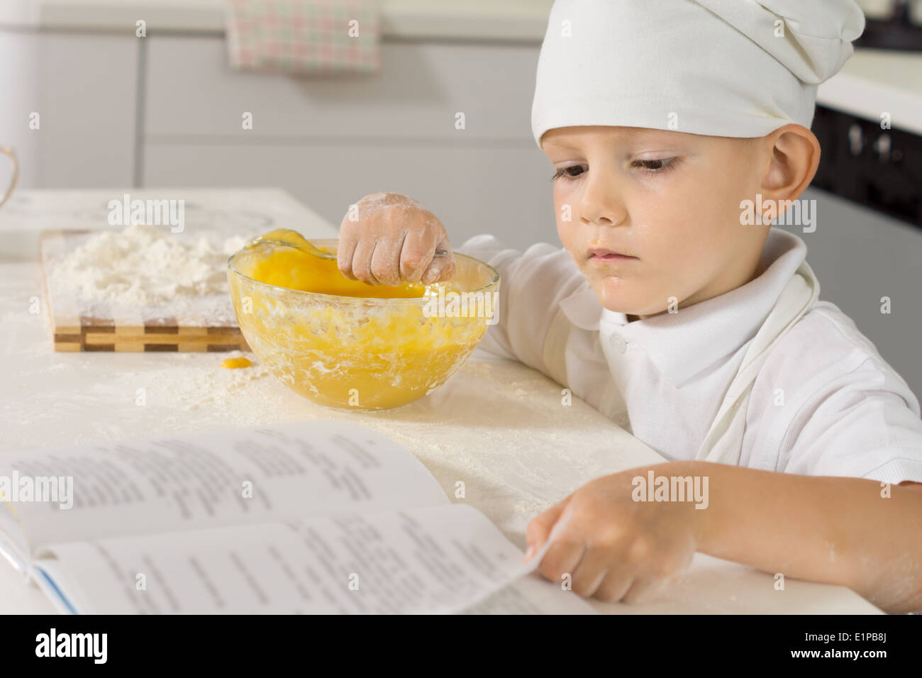 Little boy chef in a chefs uniform checking his recipe in a book as he ...