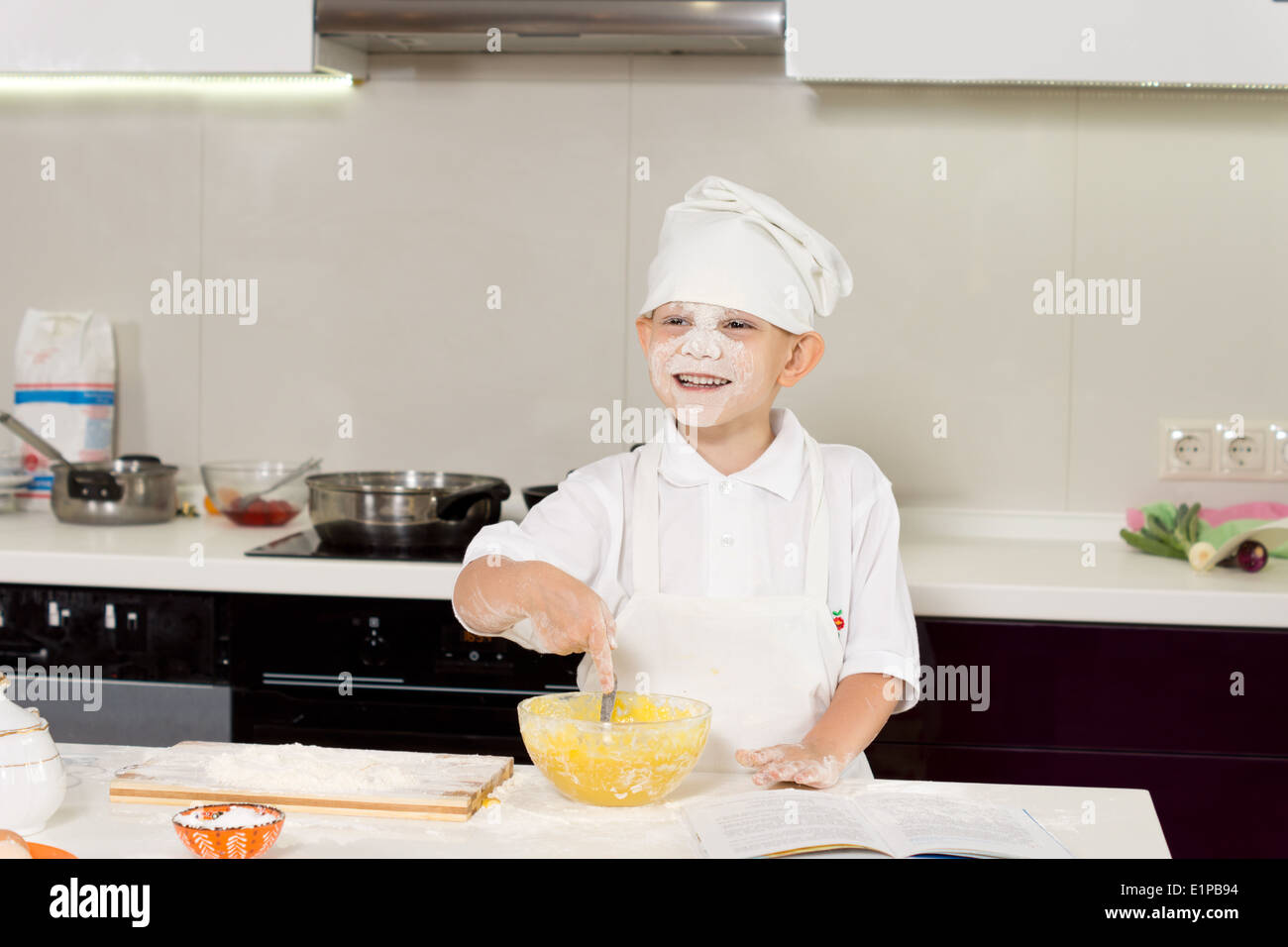 Happy cute young boy baking in the kitchen in a chefs uniform grinning ...
