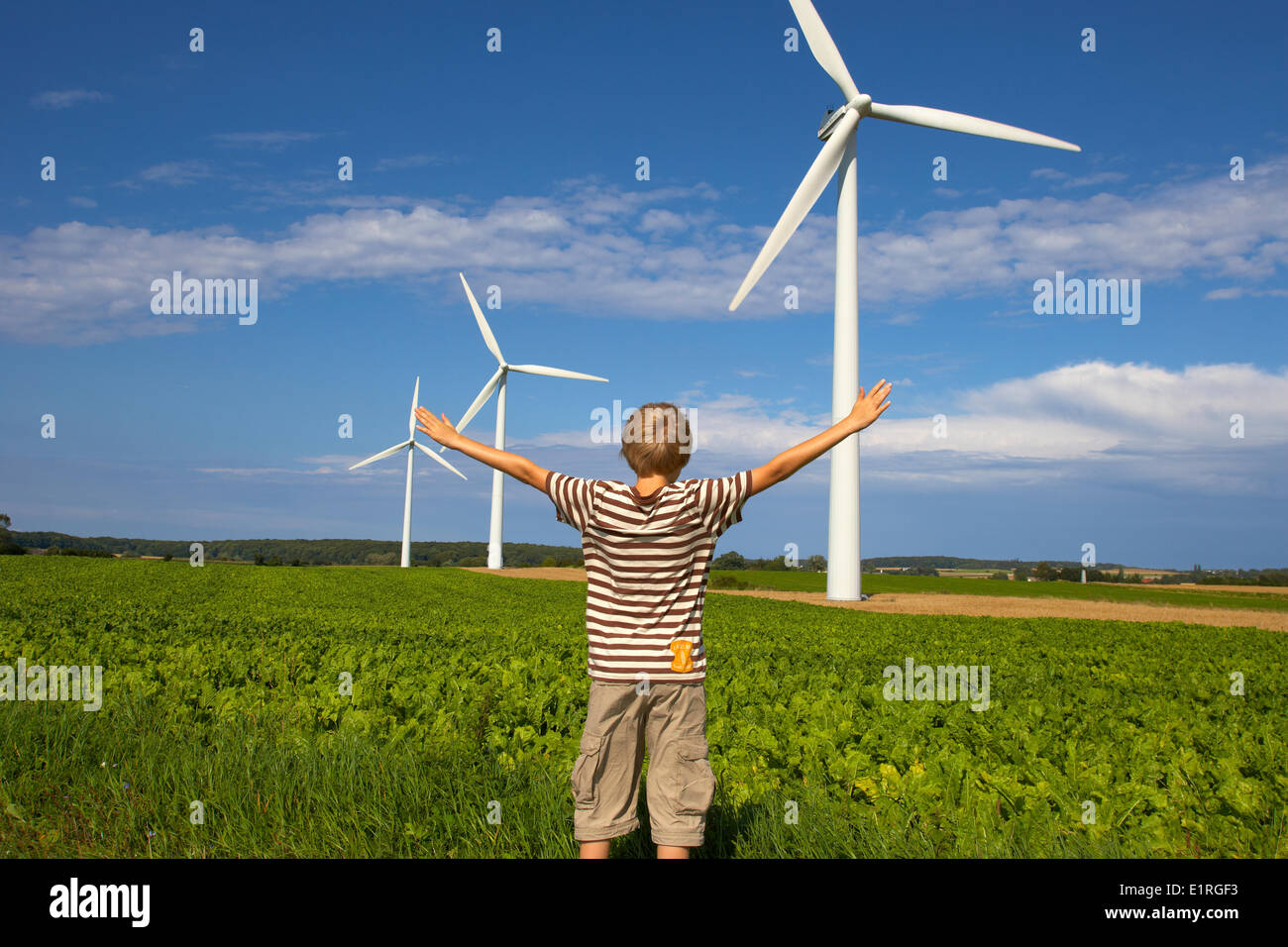 Young boy with outstreched arms facing a row of wind turbines imitating ...