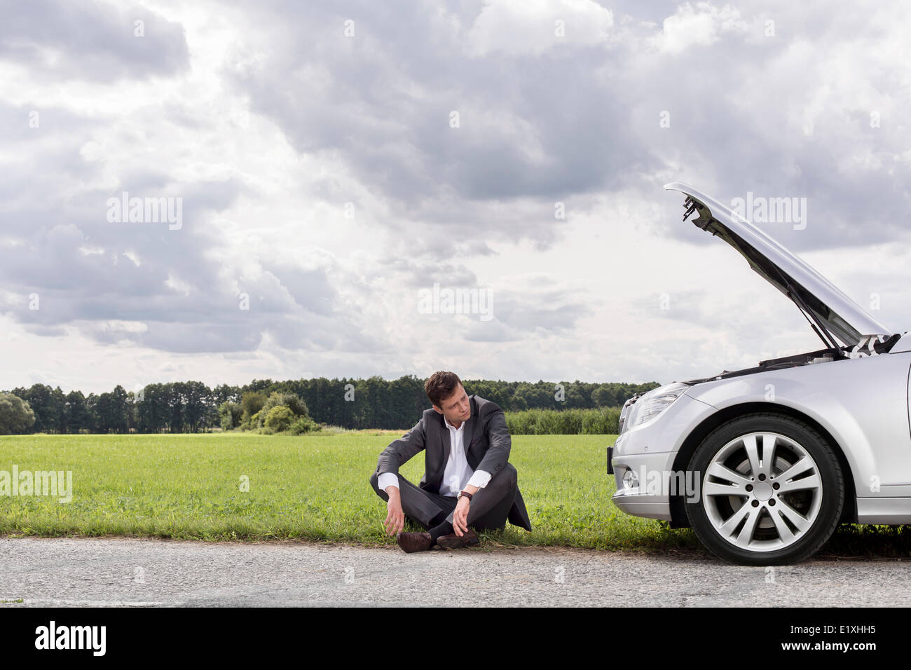 Full length of worried businessman sitting by broken down car at ...