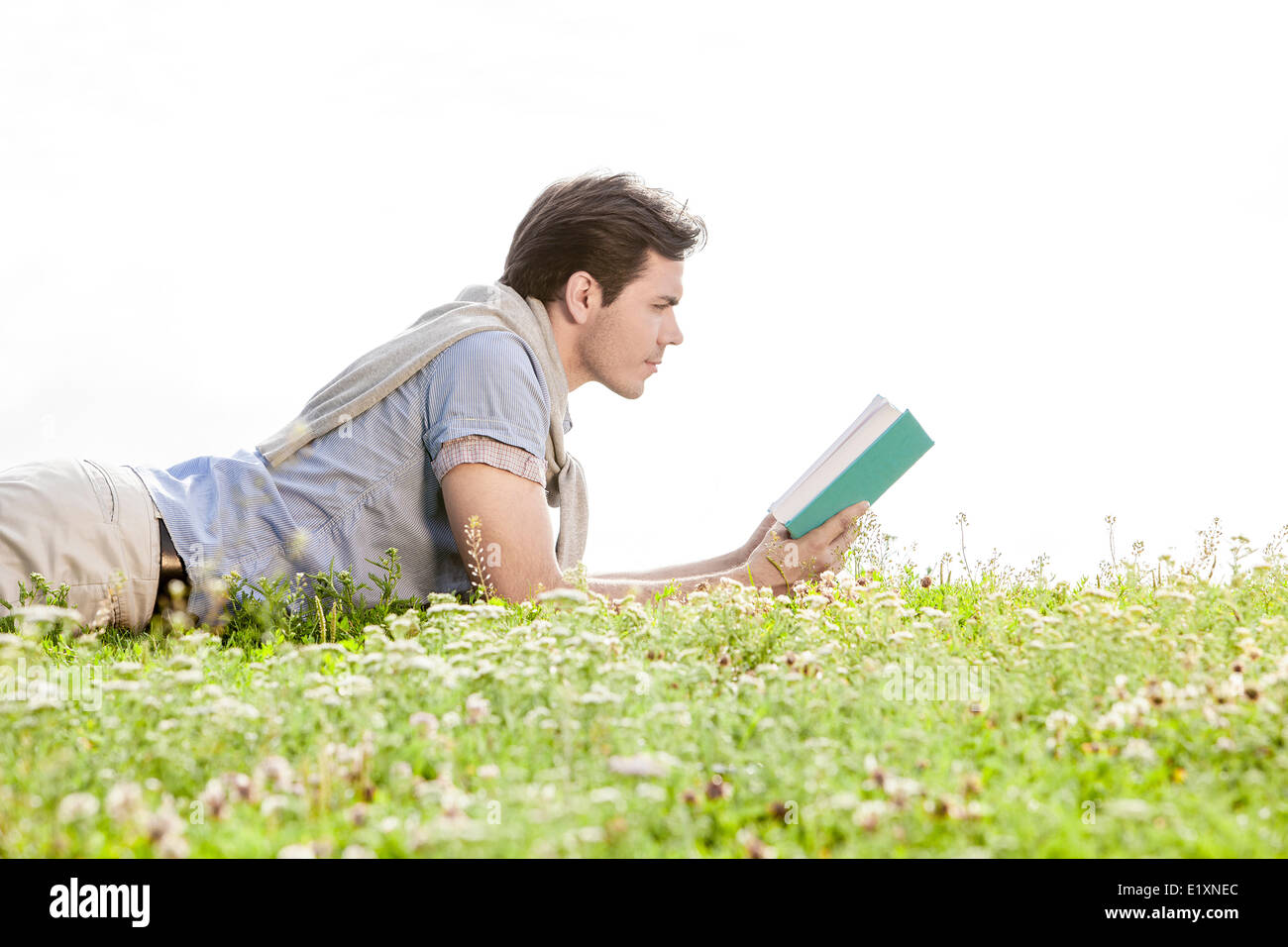 Side view of young man reading book while lying on grass against clear ...