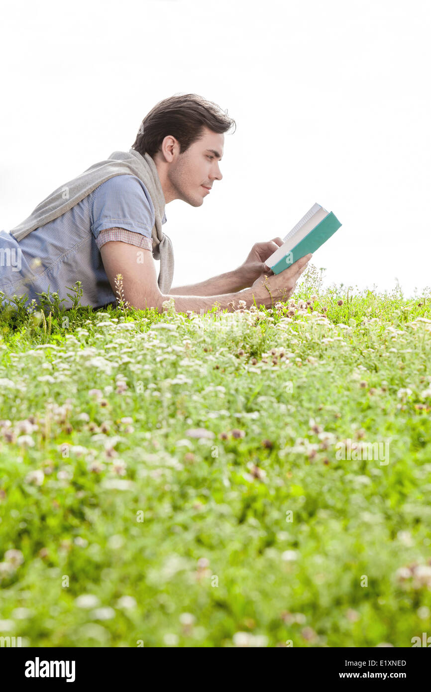 Side view of man reading book while lying on grass against clear sky ...