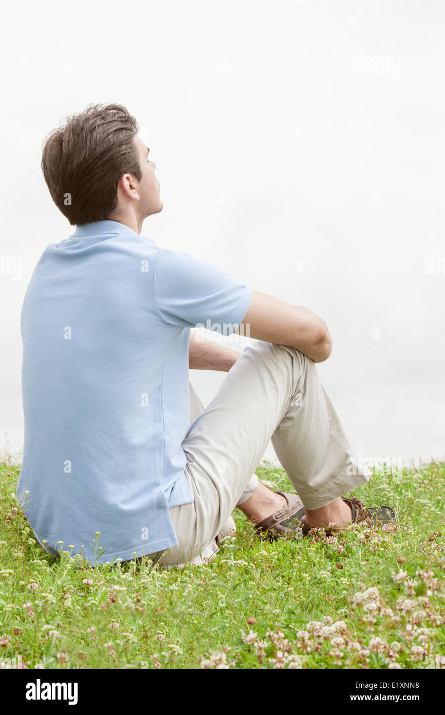 Rear view of thoughtful young man sitting on grass against sky Stock ...