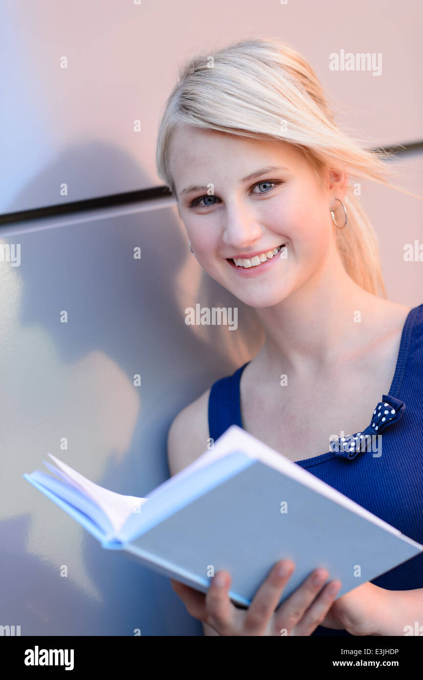 Smiling blond college student girl holding open book against wall Stock ...