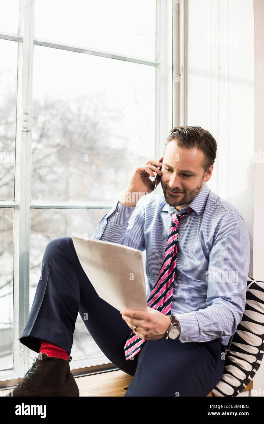 Mid adult businessman reading document while on call by office window ...
