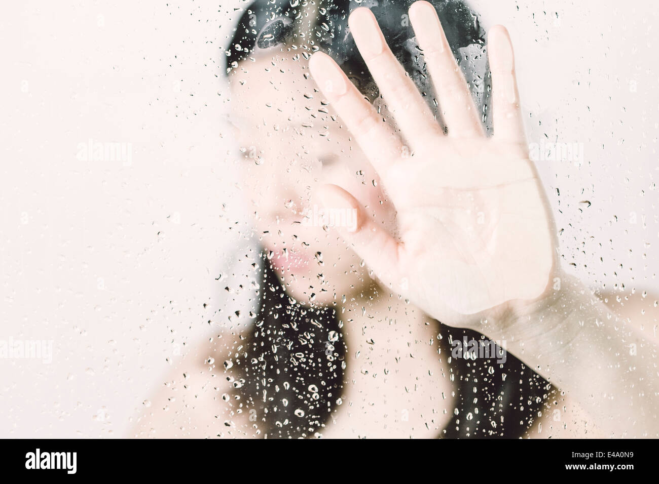 Asian woman behind window pressing hand against pane with water drops ...