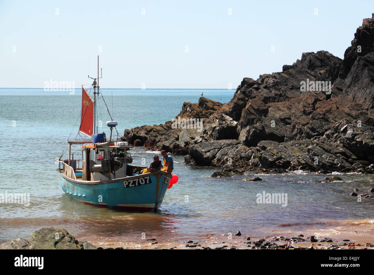 A blue fishing boat with a red mizzen sail beached on a rocky seashore ...