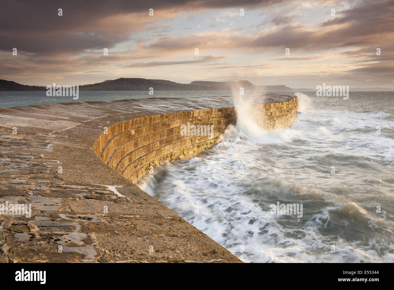 View of harbour wall with waves breaking at sunrise, looking across bay ...