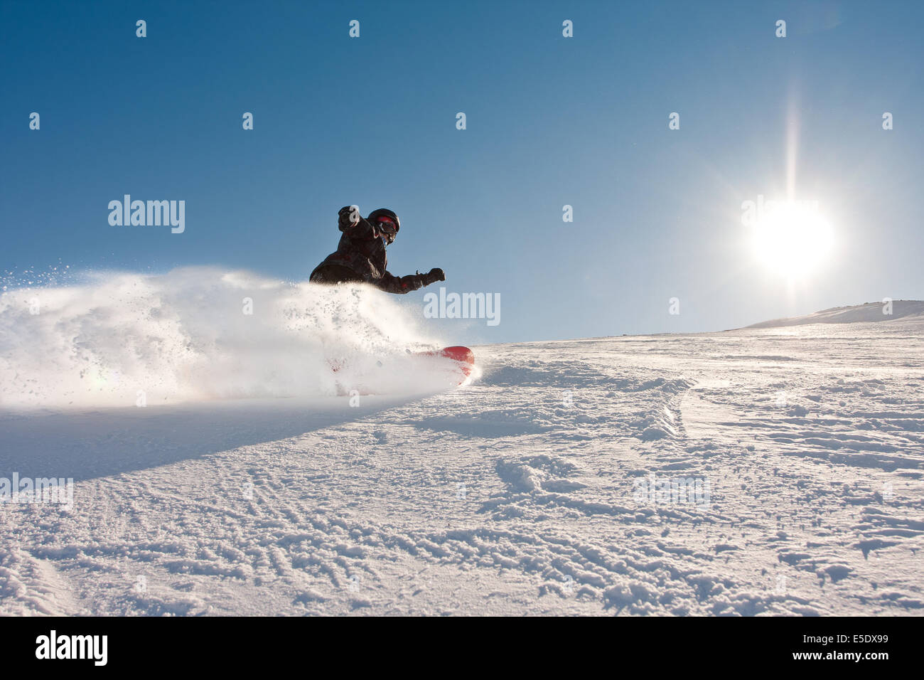 Teenage boy riding a snowboard downhill in sunny weather, spraying snow ...