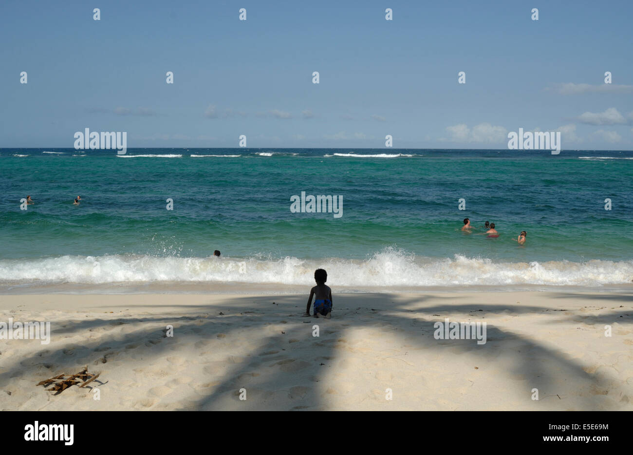 Small boy playing on beach, in the shadow formed by a palm tree. Back ...