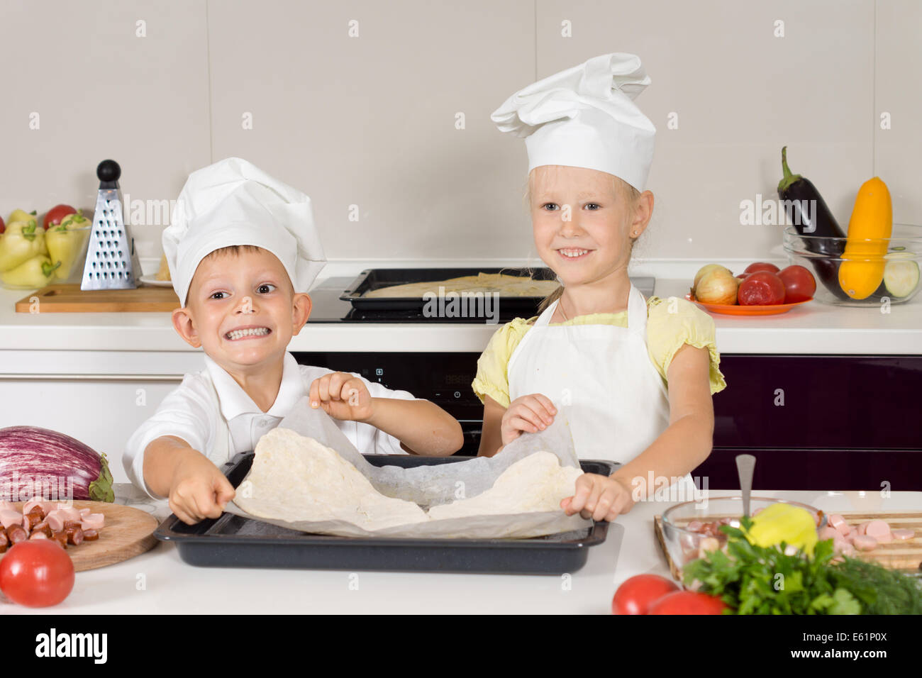 Smiling little cooks in white chefs uniforms preparing a homemade pizza ...