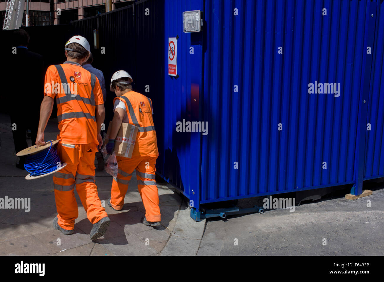Workmen in hi-visibility clothing carry a blue wiring reel ...