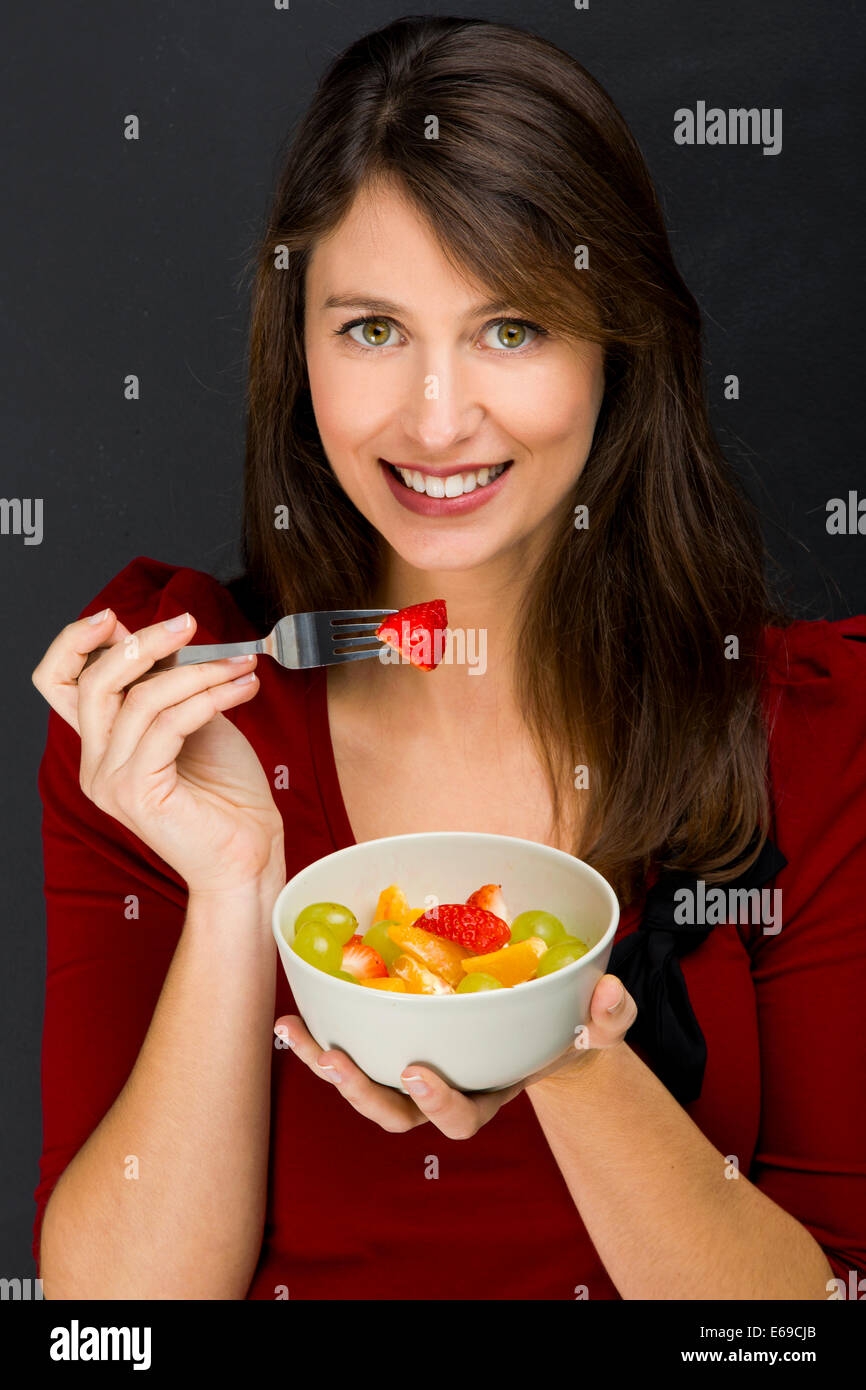 Beautiful young woman eating a fruit salad, over a black background ...