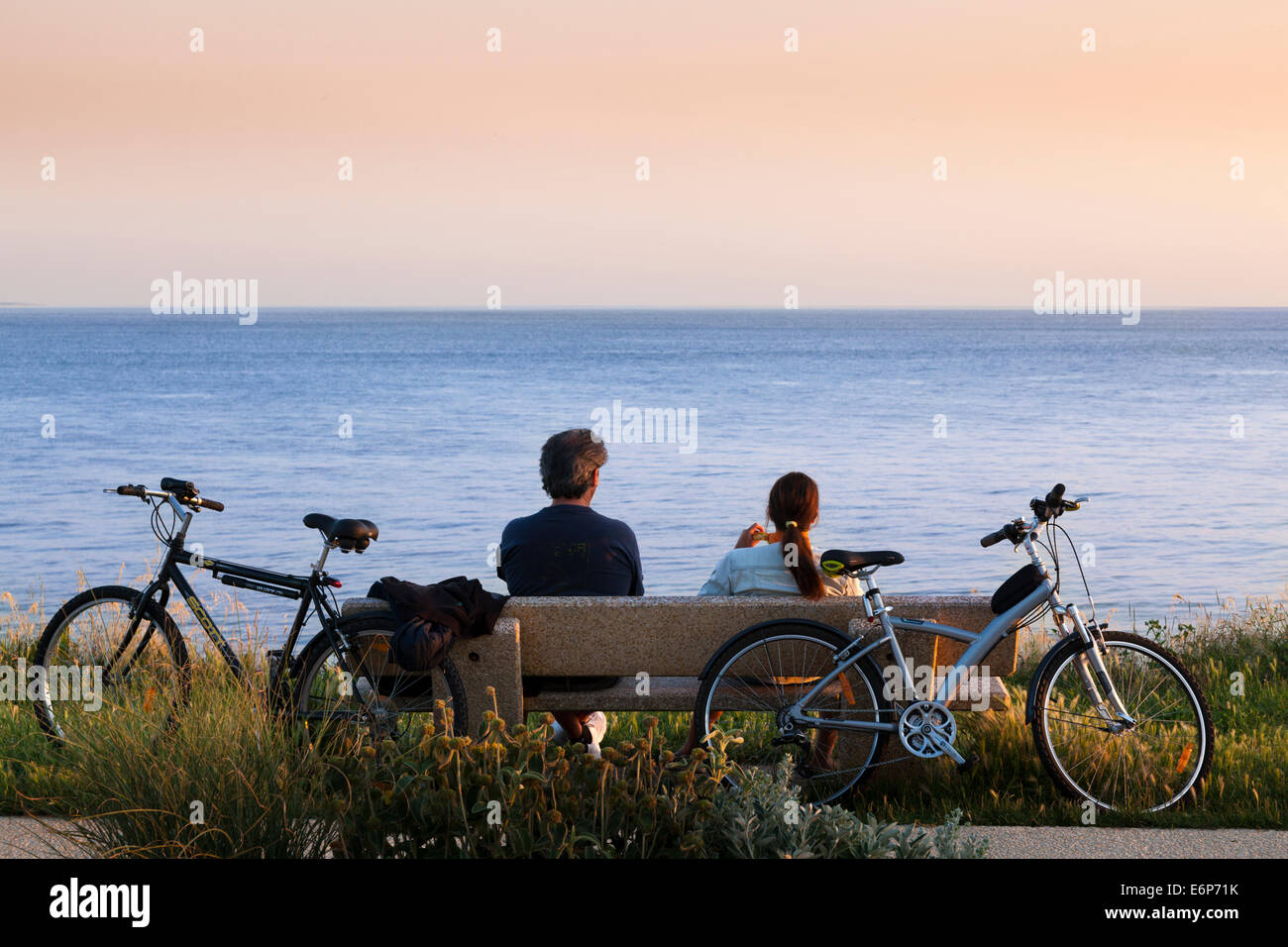 Two cyclists rest on a bench to watch the evening sky over the sea ...