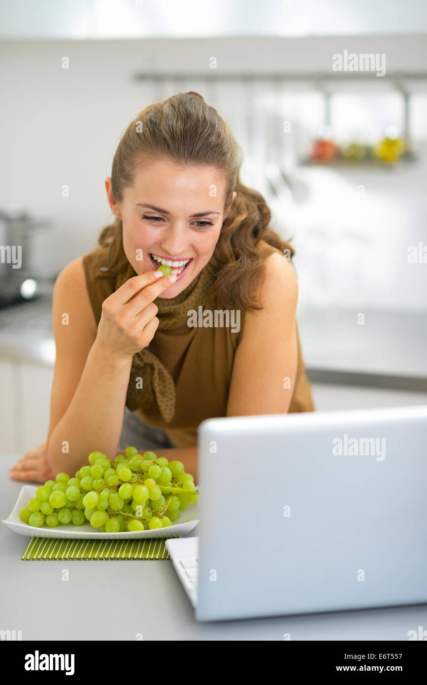 Happy young woman eating grape in kitchen and using laptop Stock Photo ...