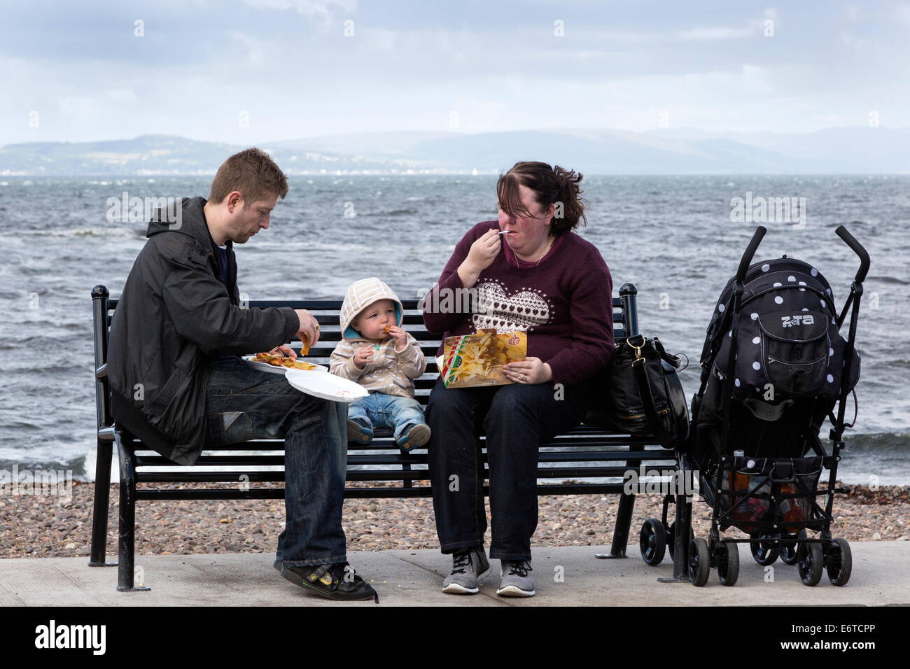 Family at the seaside, sitting on a beach bench and eating a fish ...