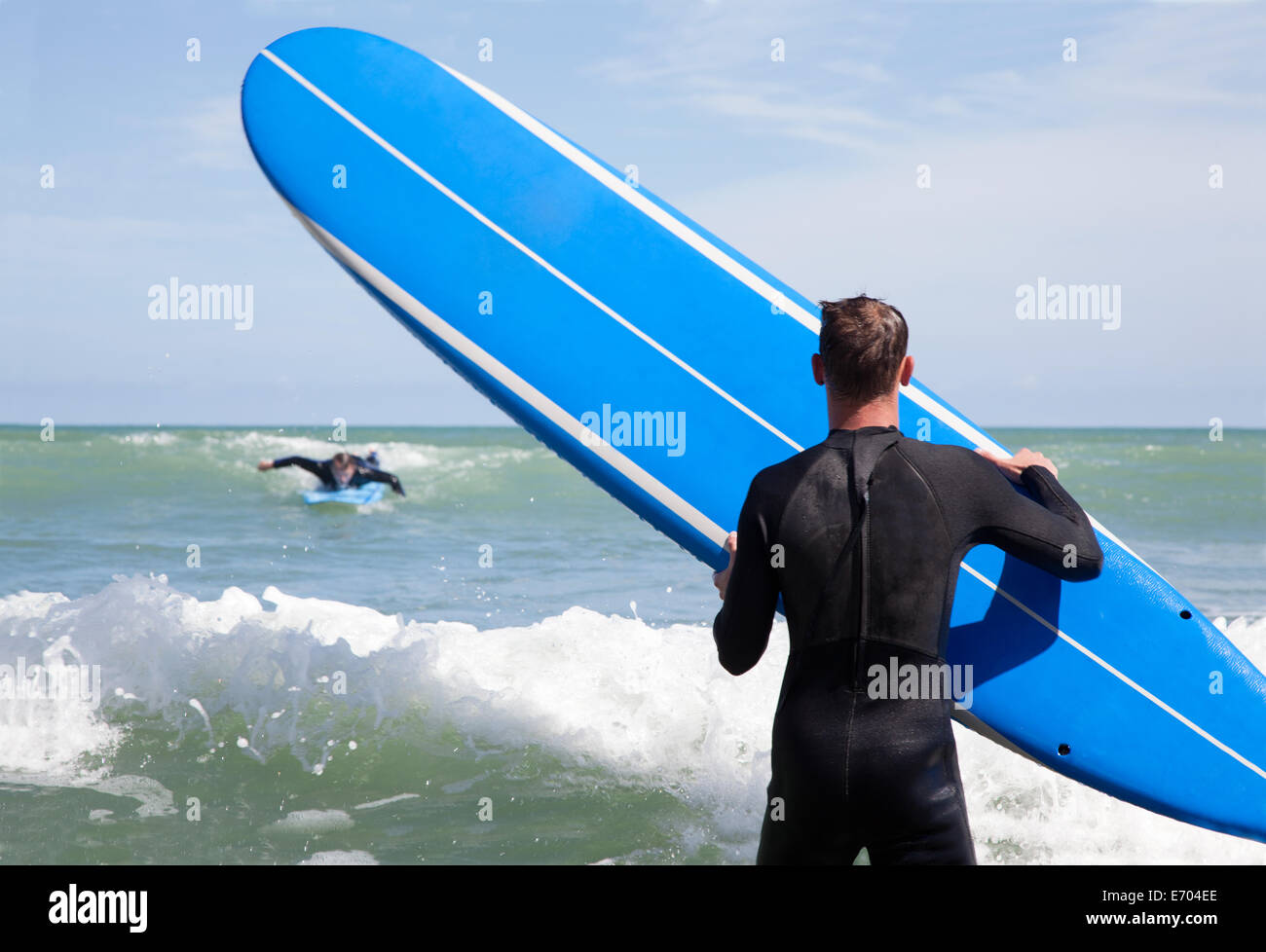 Rear view of male surfer with surfboard watching friend surfing Stock ...
