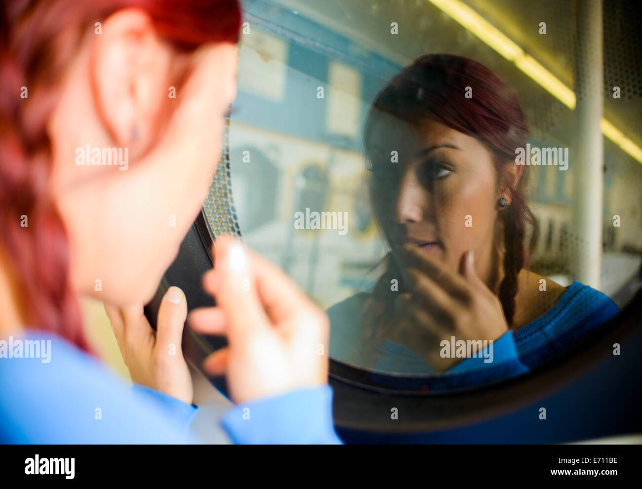 Young woman in laundromat, looking at reflection in washing machine ...