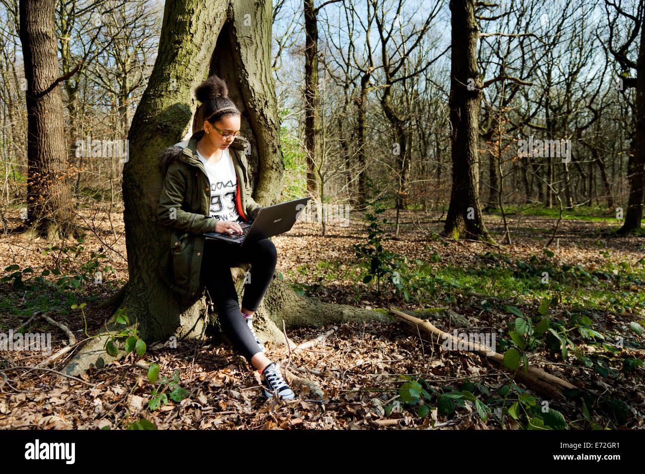 Young mixed race girl leans against a tree in a forest and logs on to ...
