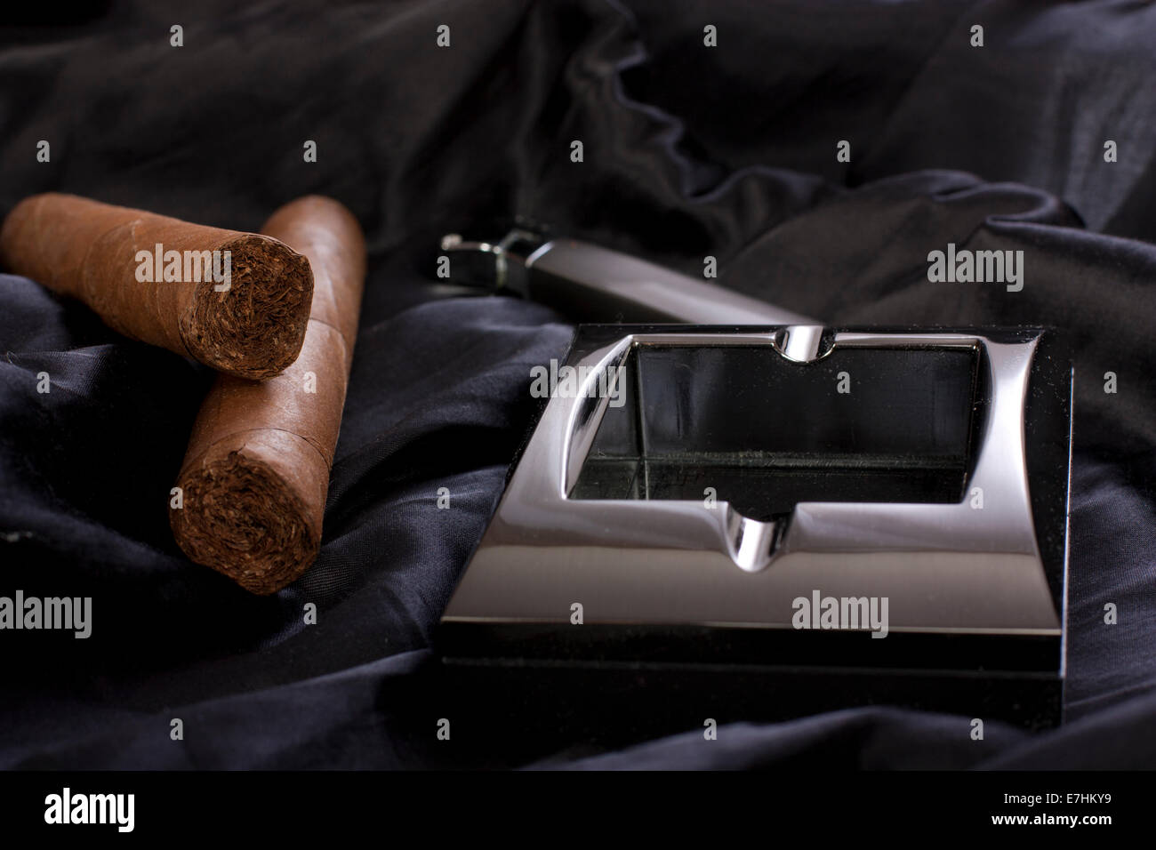 Close up of metallic ashtray, lighter and cuban cigar on black silk ...