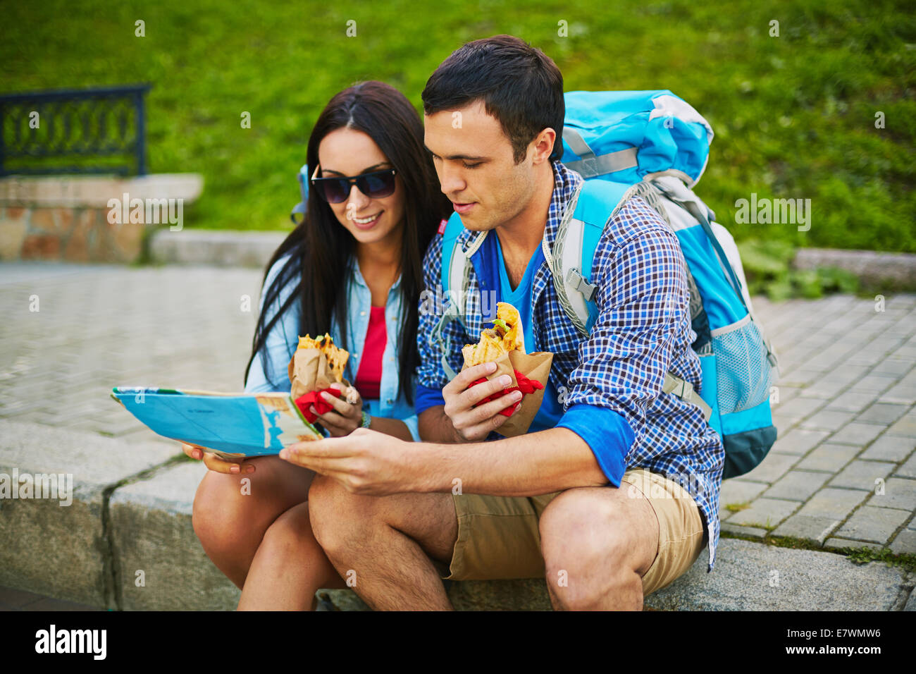 Young travelers looking at map while sitting on pavement and eating ...