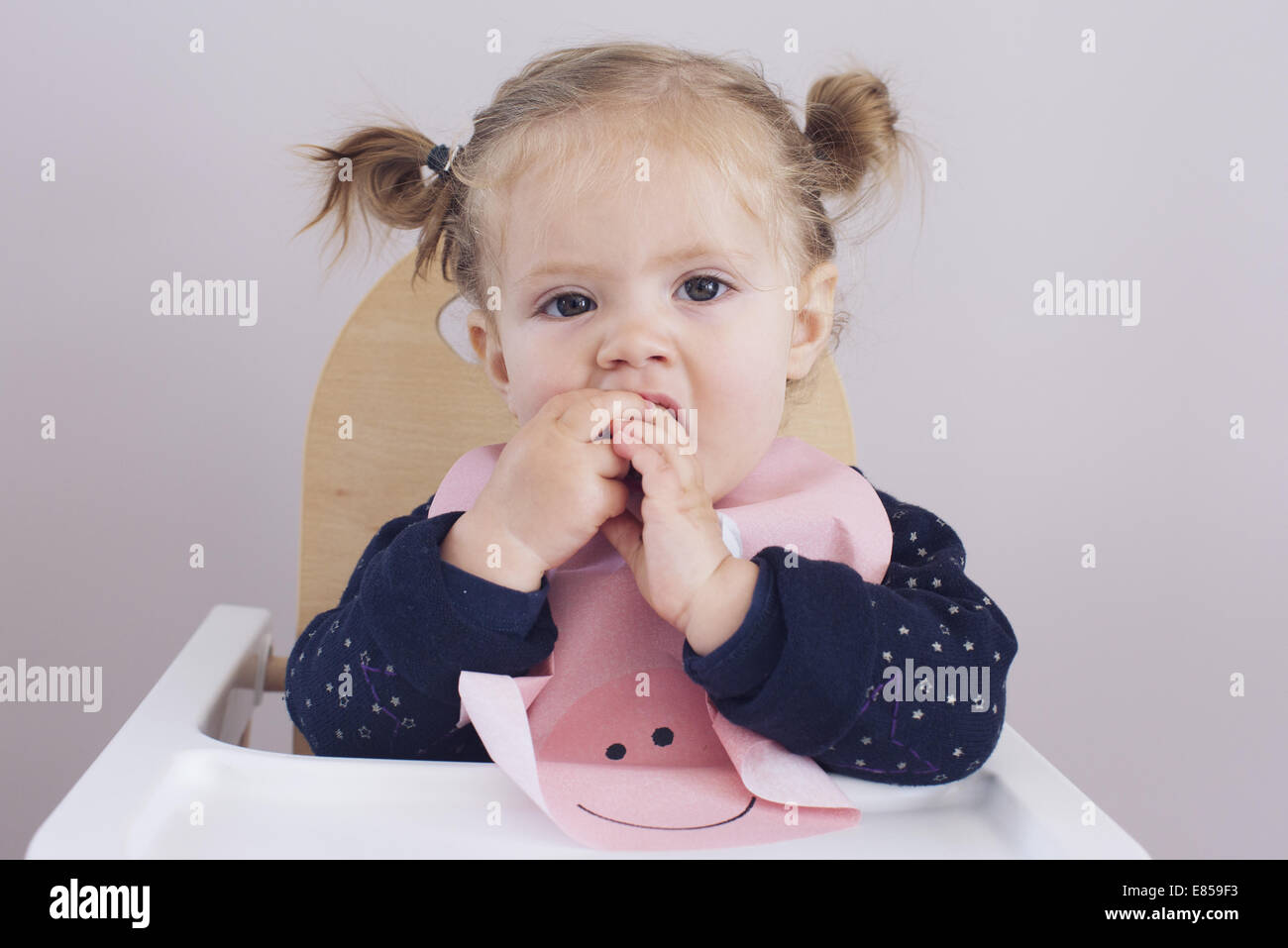 Baby girl sitting in high chair, chewing on fingers Stock Photo - Alamy