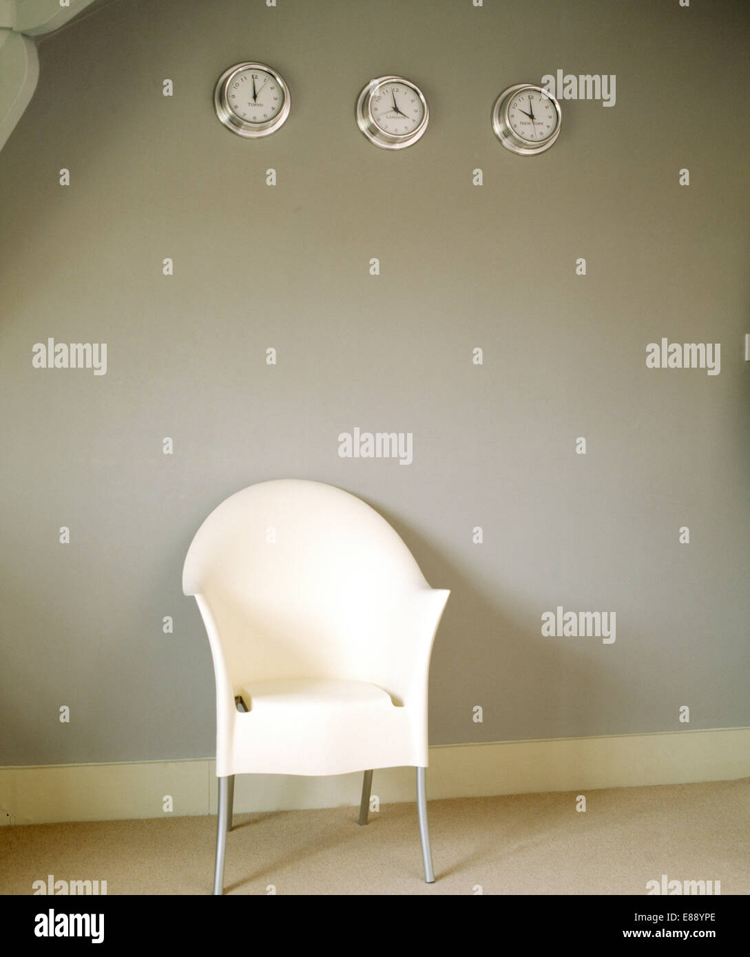 Chrome clocks on wall above white moulded plastic chair in grey hall ...