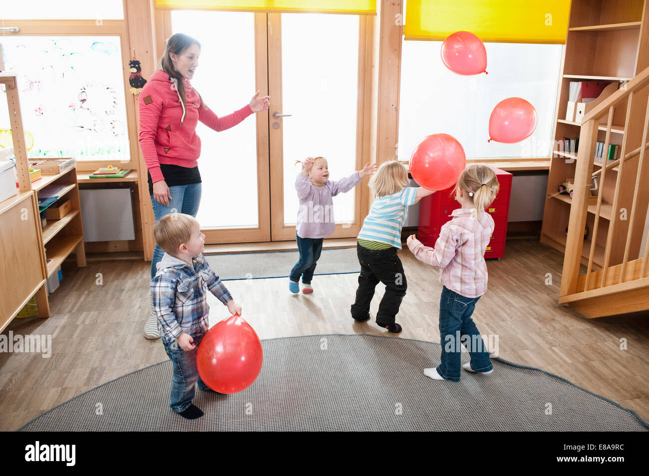 Female educator and four kids playing with red balloons in kindergarten ...