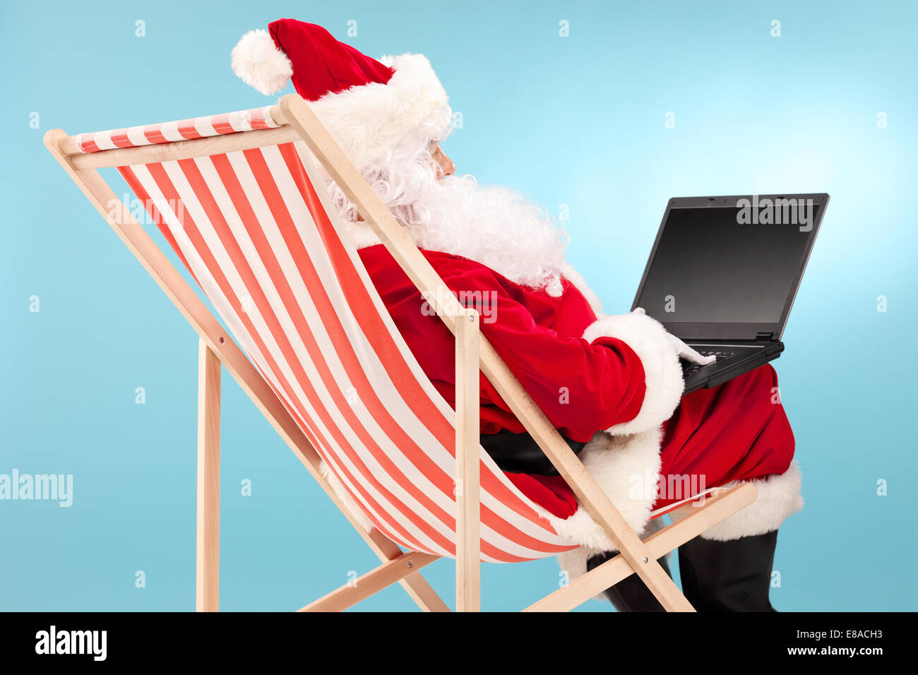 Santa Claus working on a laptop seated in a sun lounger on a blue ...