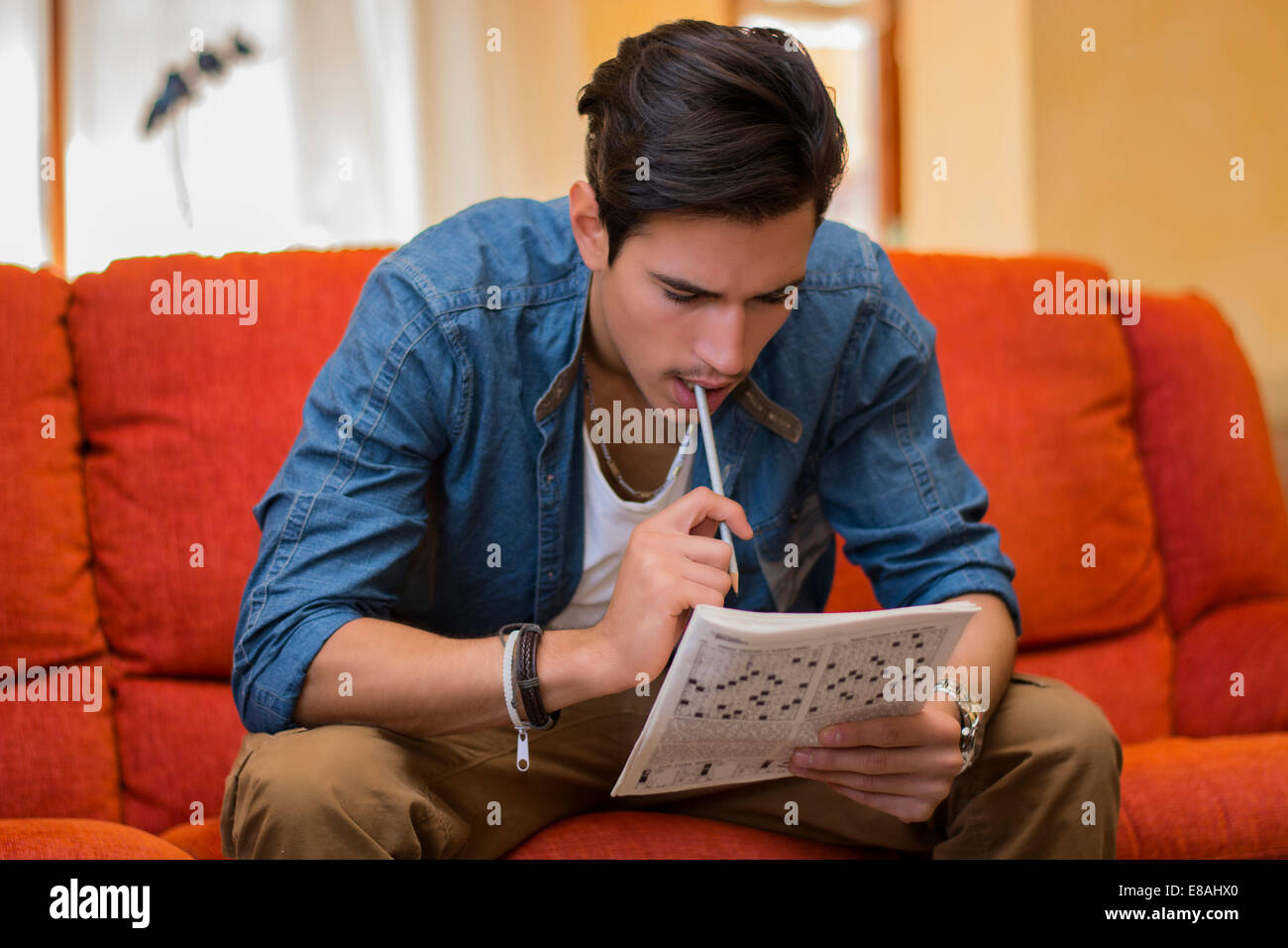 Young Man Sitting Doing A Crossword Puzzle Looking Thoughtfully At A Magazine With His Pencil To