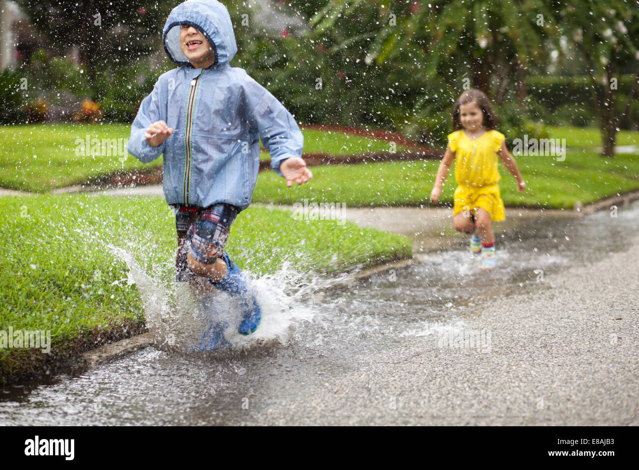 Boy and sister wearing rubber boots running and splashing in rain ...