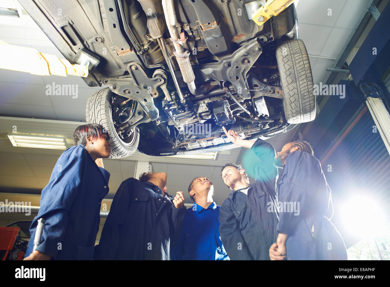 Five college students looking up from beneath car in garage workshop ...
