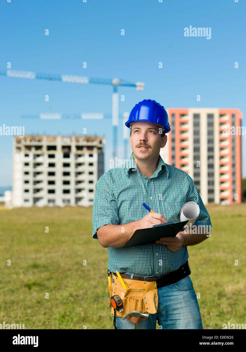 caucasian engineer standing, writing on clipboard, with buildings in ...