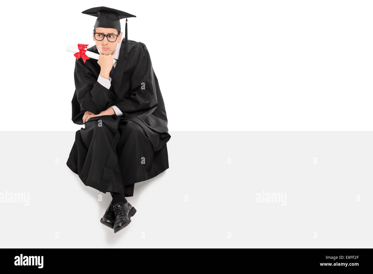 Pensive college graduate sitting on a blank panel isolated on white ...