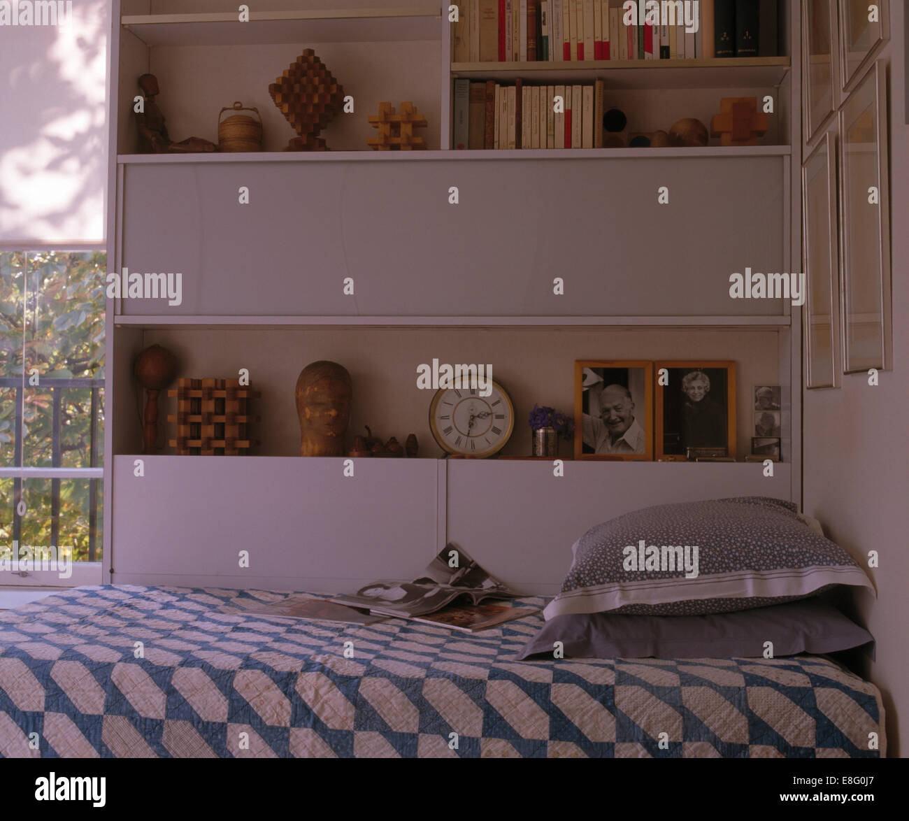 Alarm clock and books on shelves beside bed with blue+white quilt and ...