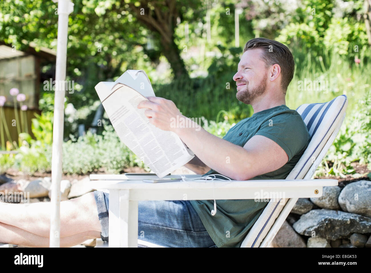 Side view of smiling man reading newspaper on lounge chair at yard ...
