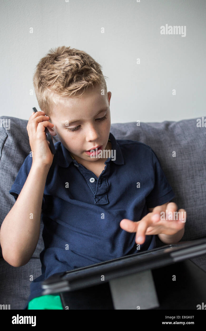 Boy answering smart phone while using digital tablet on sofa at home ...