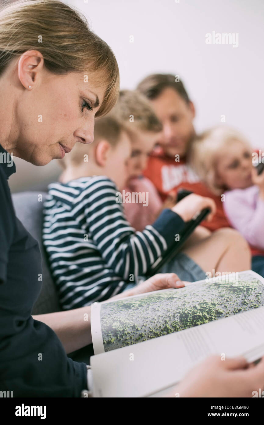 Side view of woman reading magazine with family using technologies in ...