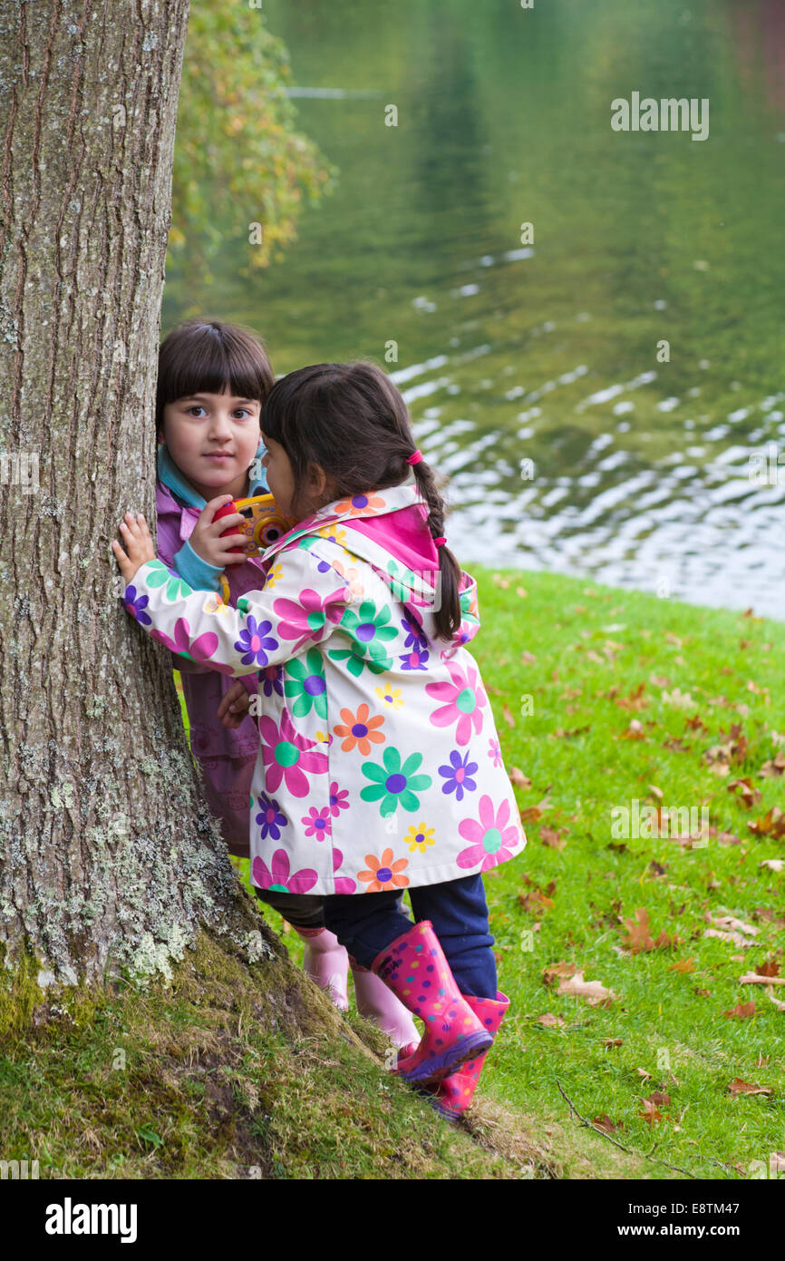 Two young girls, one with toy camera, playing around tree in October ...