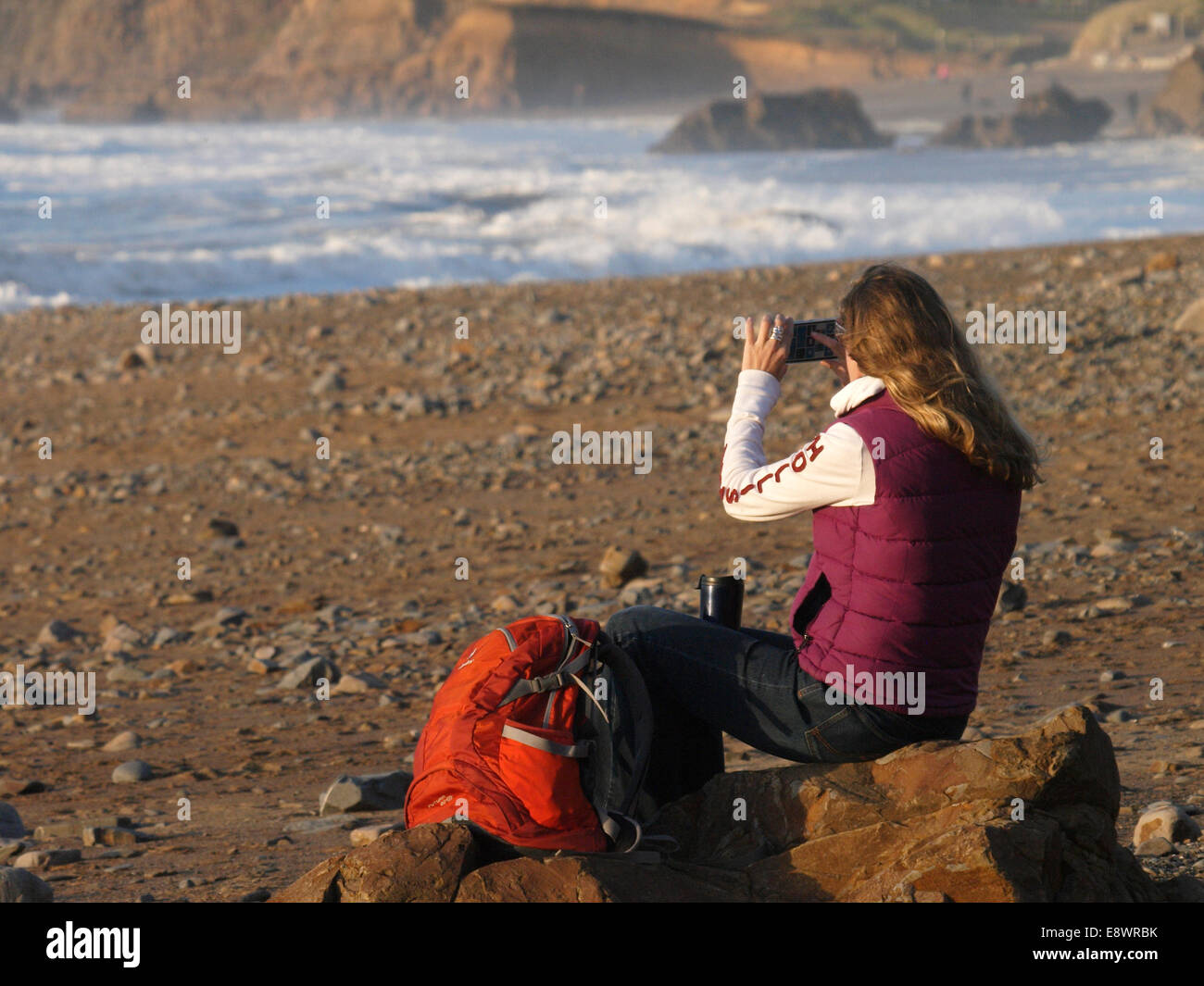 Woman sat on a rock at the beach taking photo with mobile phone ...