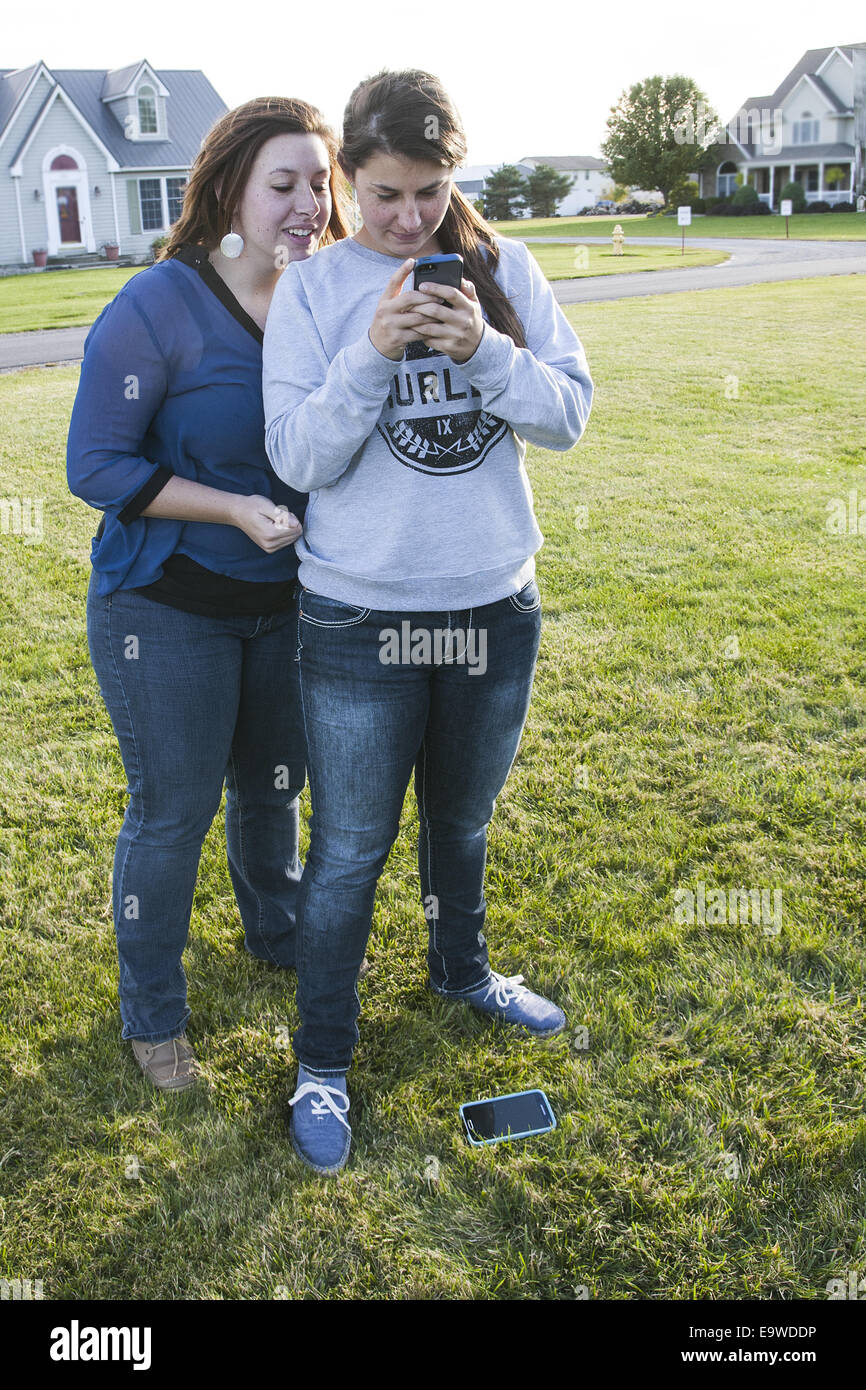 two 20-30's women are standing outside looking closely at a cell phone ...