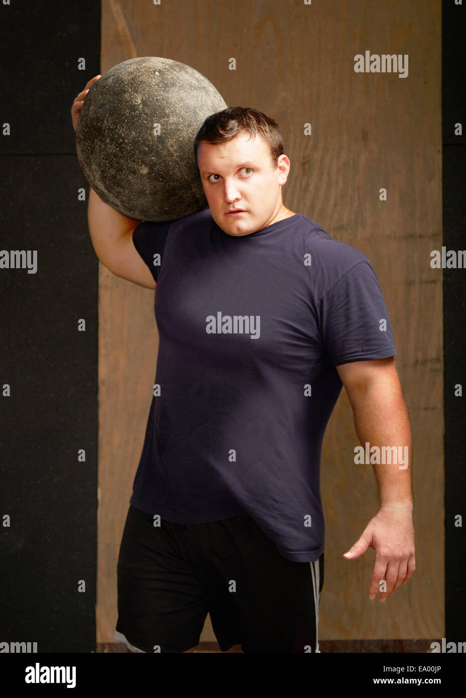 Portrait of young male weightlifter with atlas ball on shoulder in gym ...