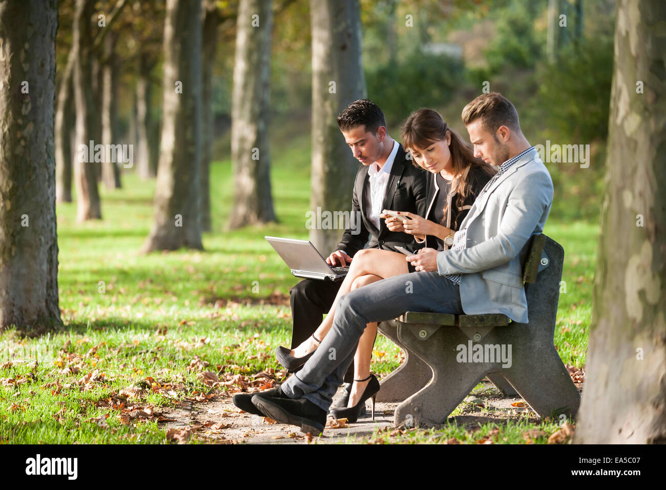 Three business people sitting on a park bench using laptop, smartphone ...