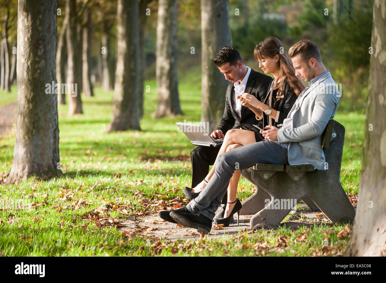 Three business people sitting on a park bench using laptop, smartphone ...