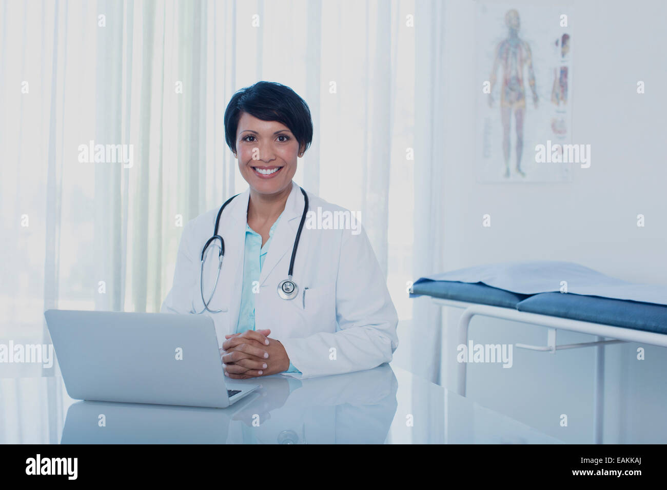 Portrait of smiling female doctor sitting at desk with laptop in office ...