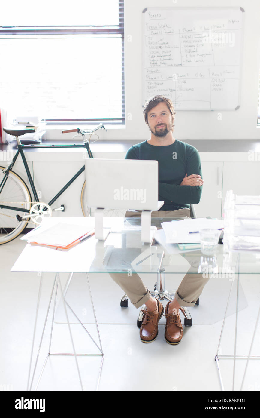 Portrait of man sitting with arms crossed behind glass desk in modern ...